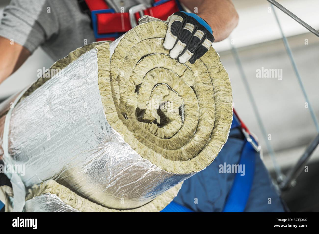 A construction worker holds rolled insulation material, preparing for installation. Safety gear ...