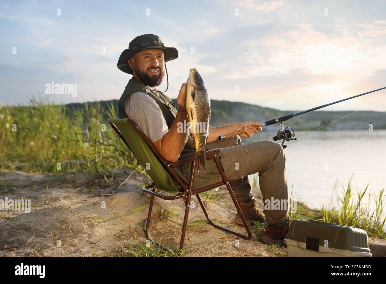 Smiling fisherman holding rod and caught fish near lake at summer Stock ...