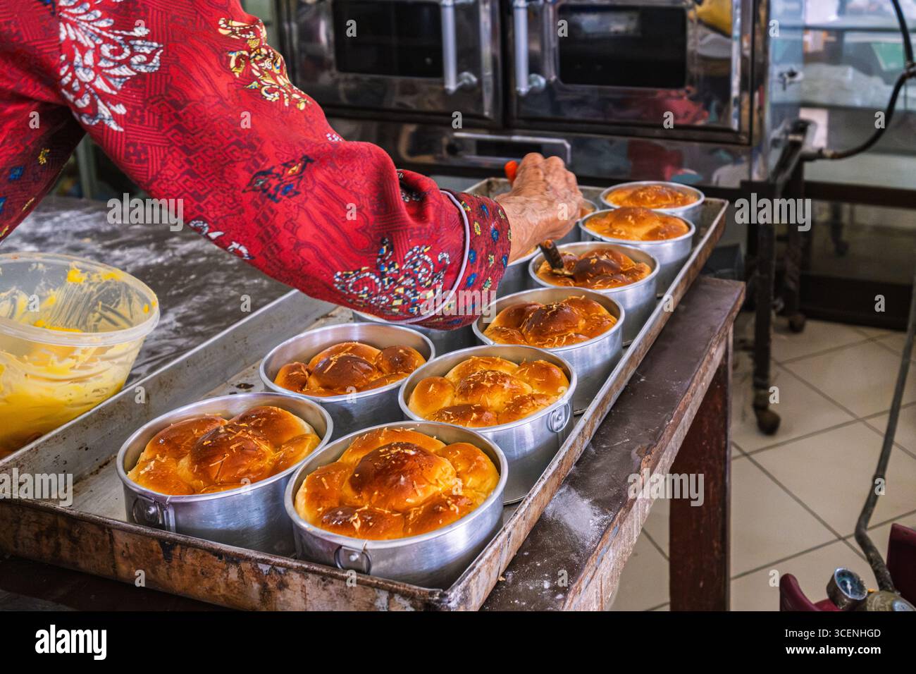 Elderly baker in red patterned attire brushing butter over golden bread ...