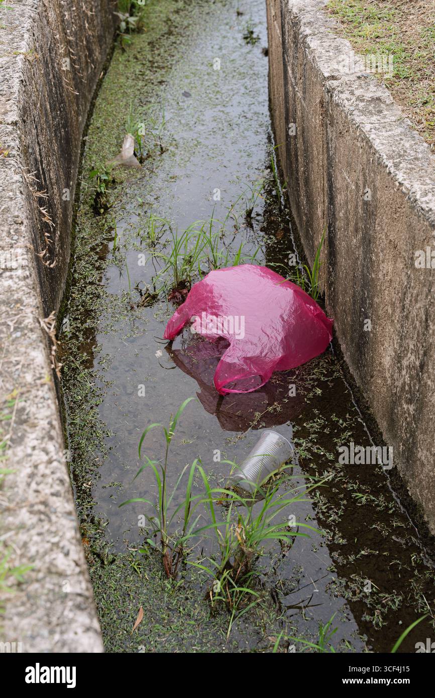A red plastic bag and other trash floating in a dirty water drainage ...