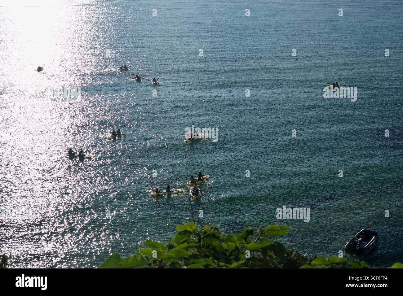 Groups of kayakers paddle across sun‑sparkled water near people in ...