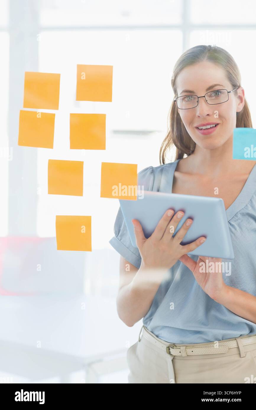 Woman in blouse standing behind sticky notes covered glass using tablet ...