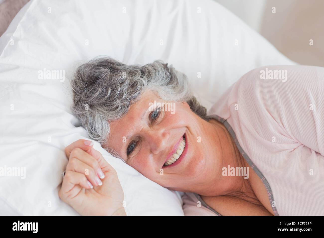 Senior woman reclining in bed in bedroom wearing pink sleepwear with ...
