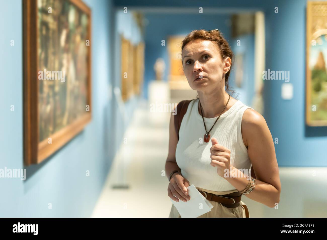 Focused woman visitor holds booklet with an exhibition program while exploring artworks in ...