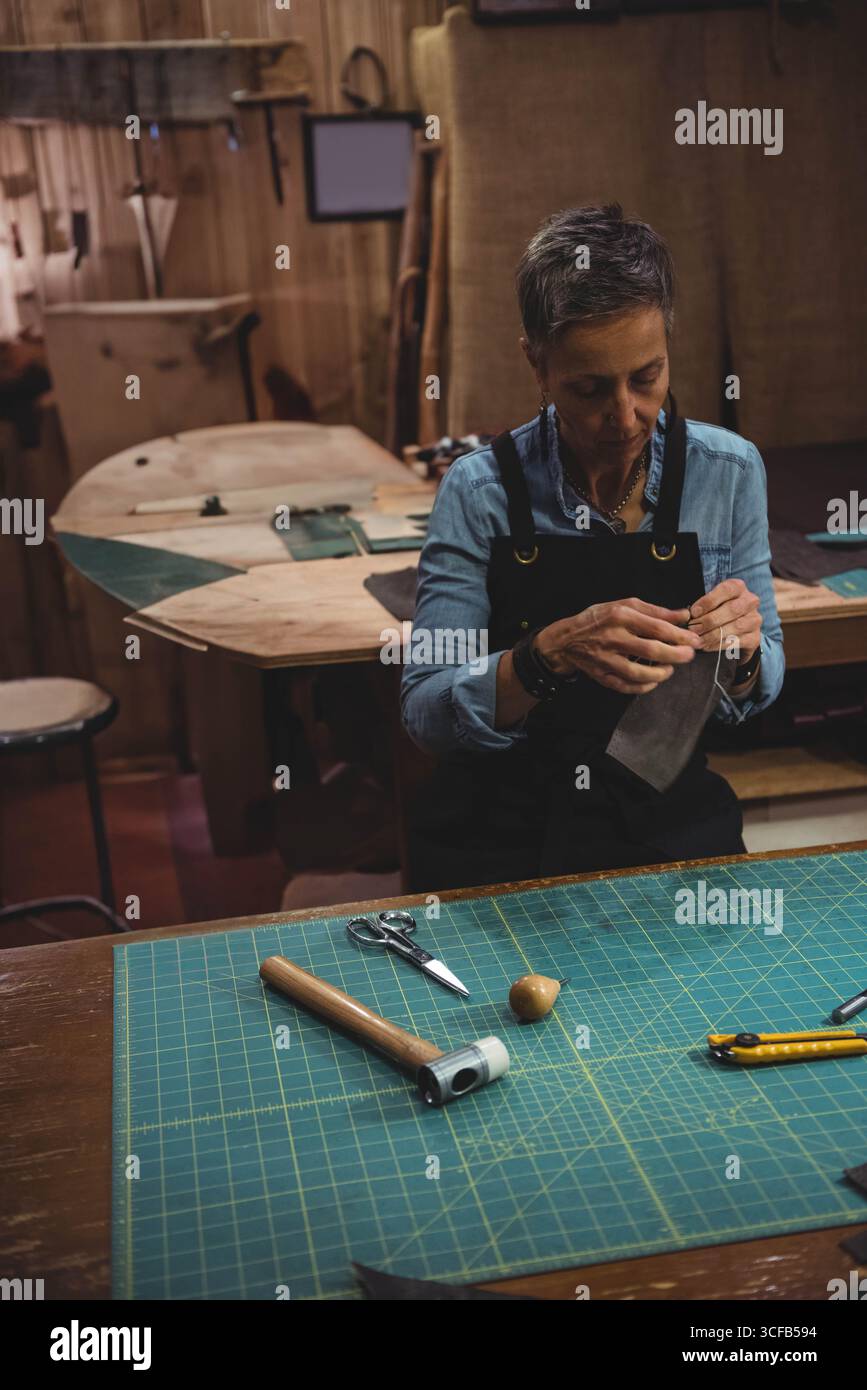 Senior woman wearing apron stitching leather by hand at workshop bench ...