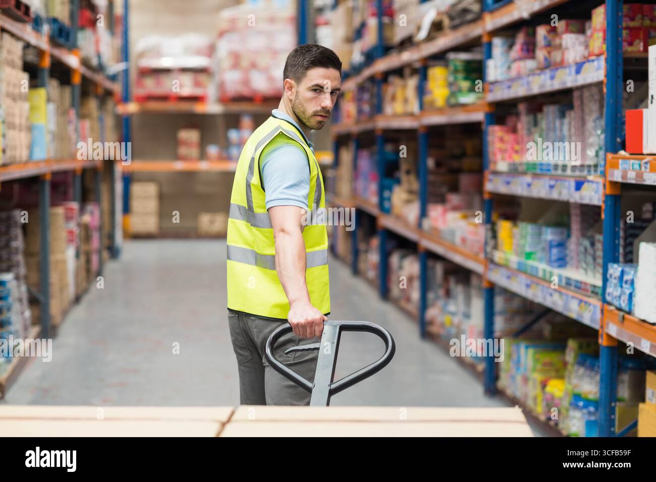 Man wearing safety vest pushing manual pallet jack along warehouse aisle with metal racks and ...