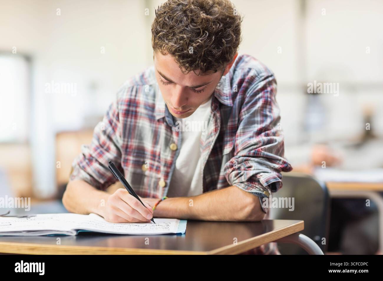 Male student writing in notebook at school desk in classroom with pen ...