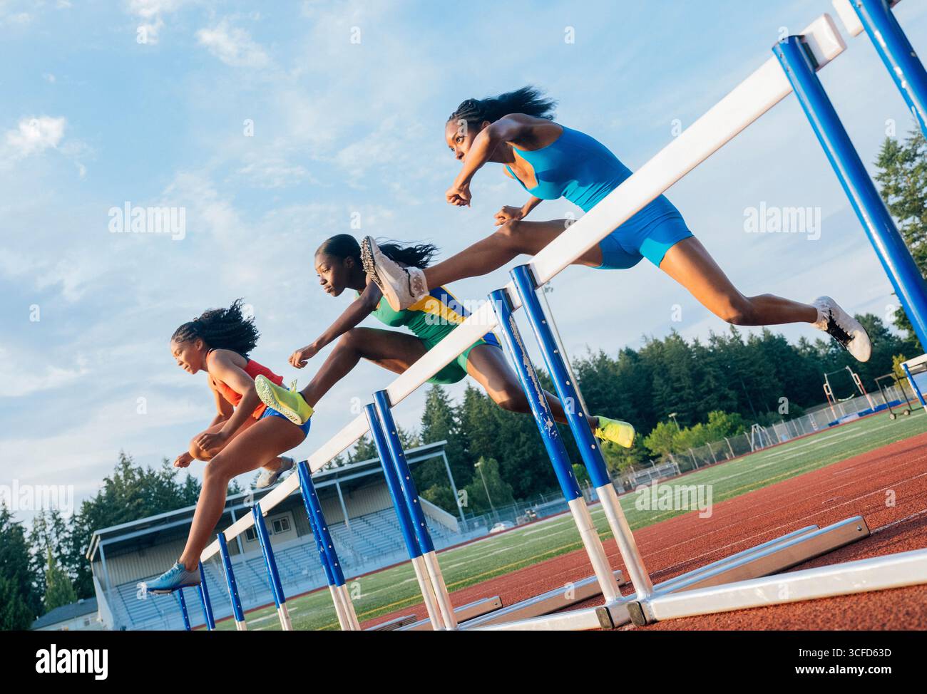 Three athletes jumping hurdles on a track under a clear blue sky during ...