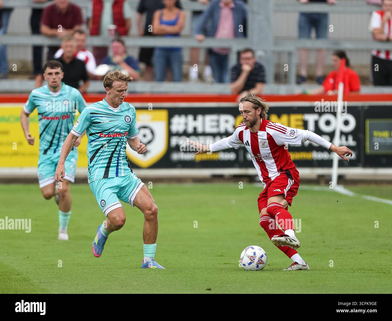 Brackley, UK. 23rd August 2025. Morgan Roberts of Brackley Town tries to beat Jack Griffiths of ...