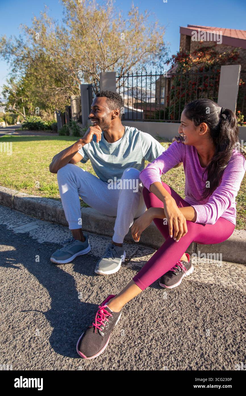 Diverse friends wearing sportswear sitting on street curb chatting under tree, metal fence Stock ...