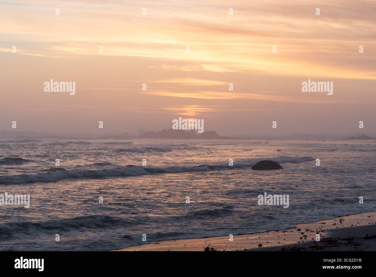 The sun sinks behind ocean waves as scattered rocks and warm light ...