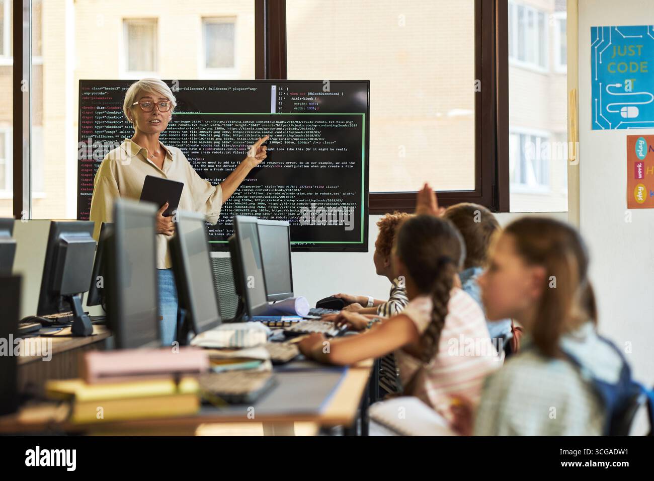Senior Caucasian woman teaching group of diverse children coding in computer classroom, standing ...