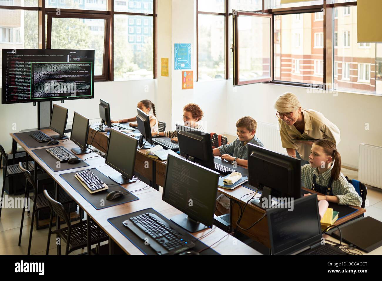 Caucasian female teacher assisting group of children using desktop computers in classroom ...