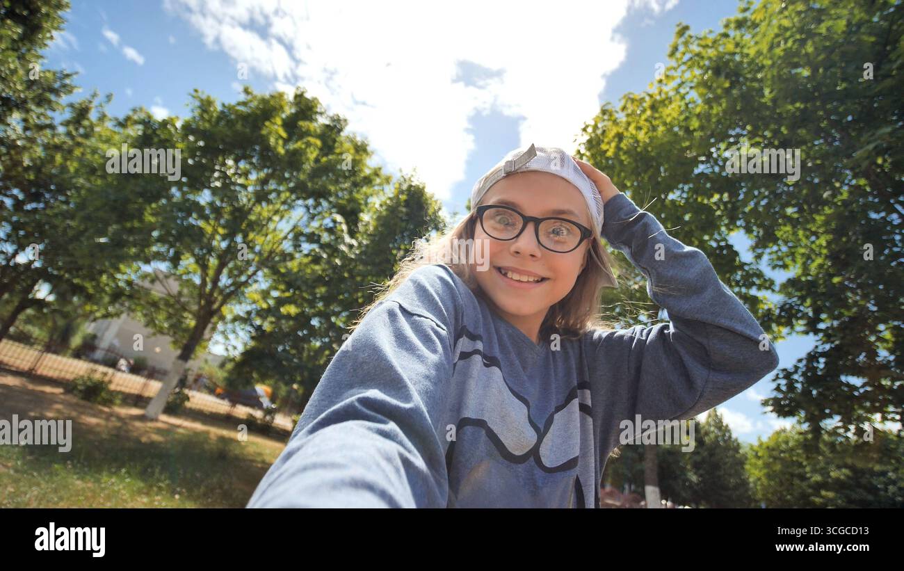 Schoolgirl taking selfie outdoors, wearing glasses, cap, surrounded by ...
