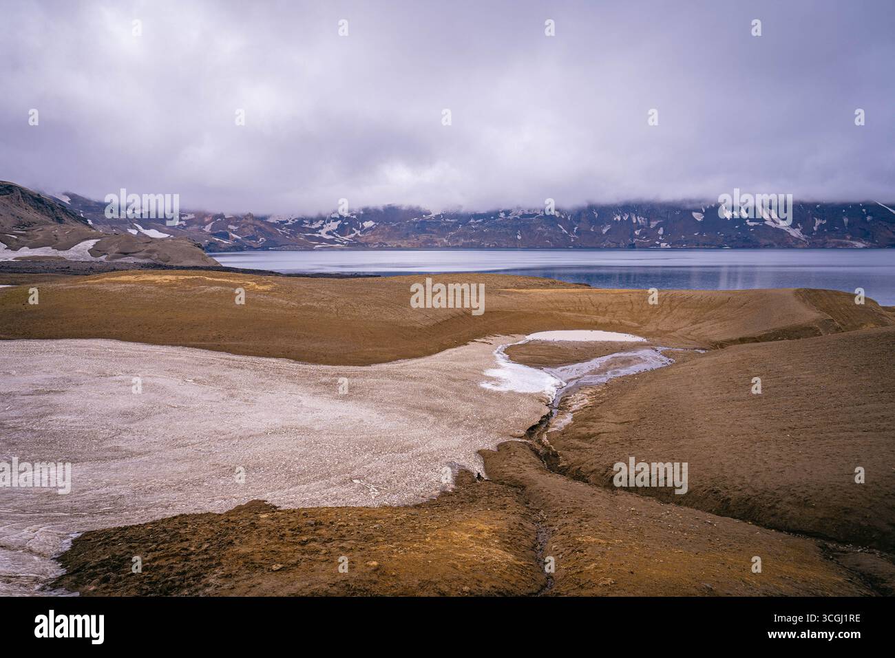 Snow patches and volcanic earth frame the shoreline of Iceland’s Askja ...