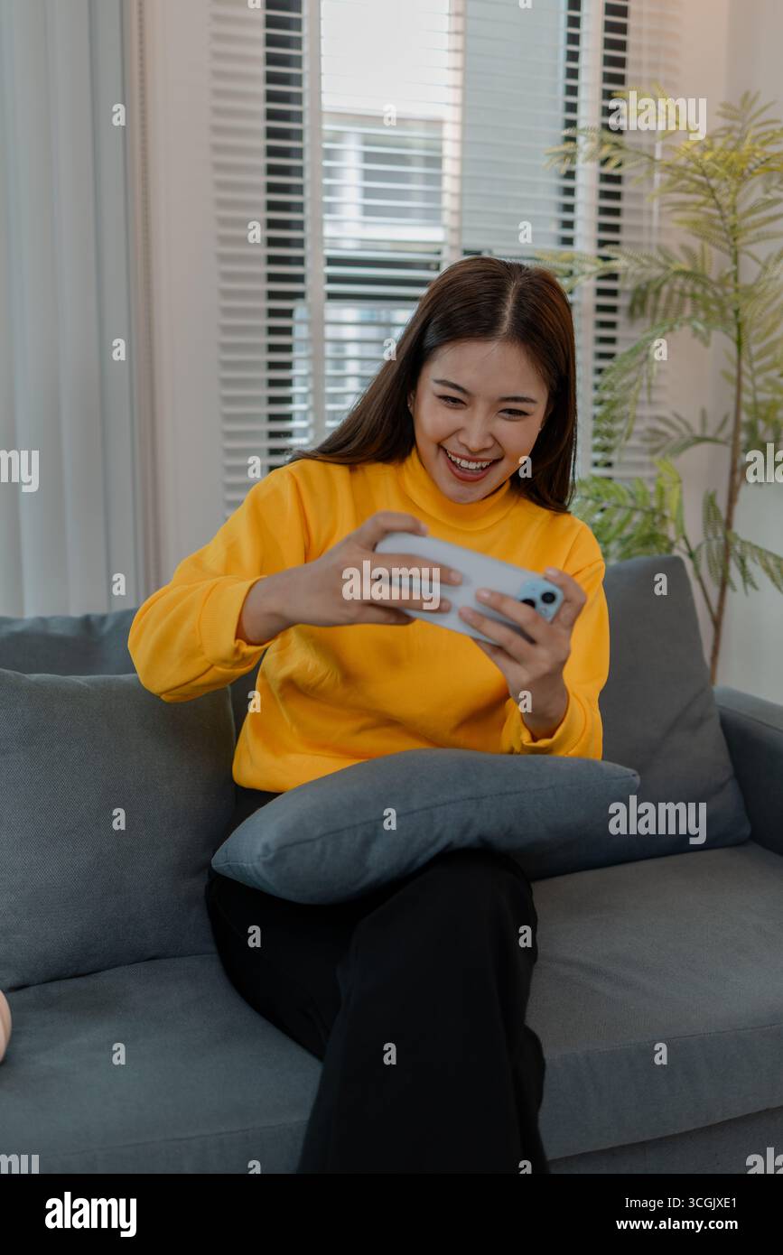 Young Asian woman in yellow sweatshirt sitting on sofa at home ...