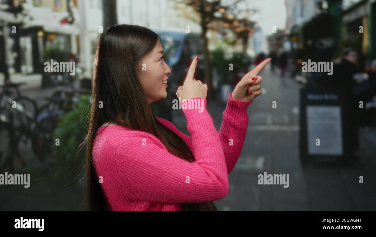 Woman smiling in pink sweater stands on city street pointing upwards ...