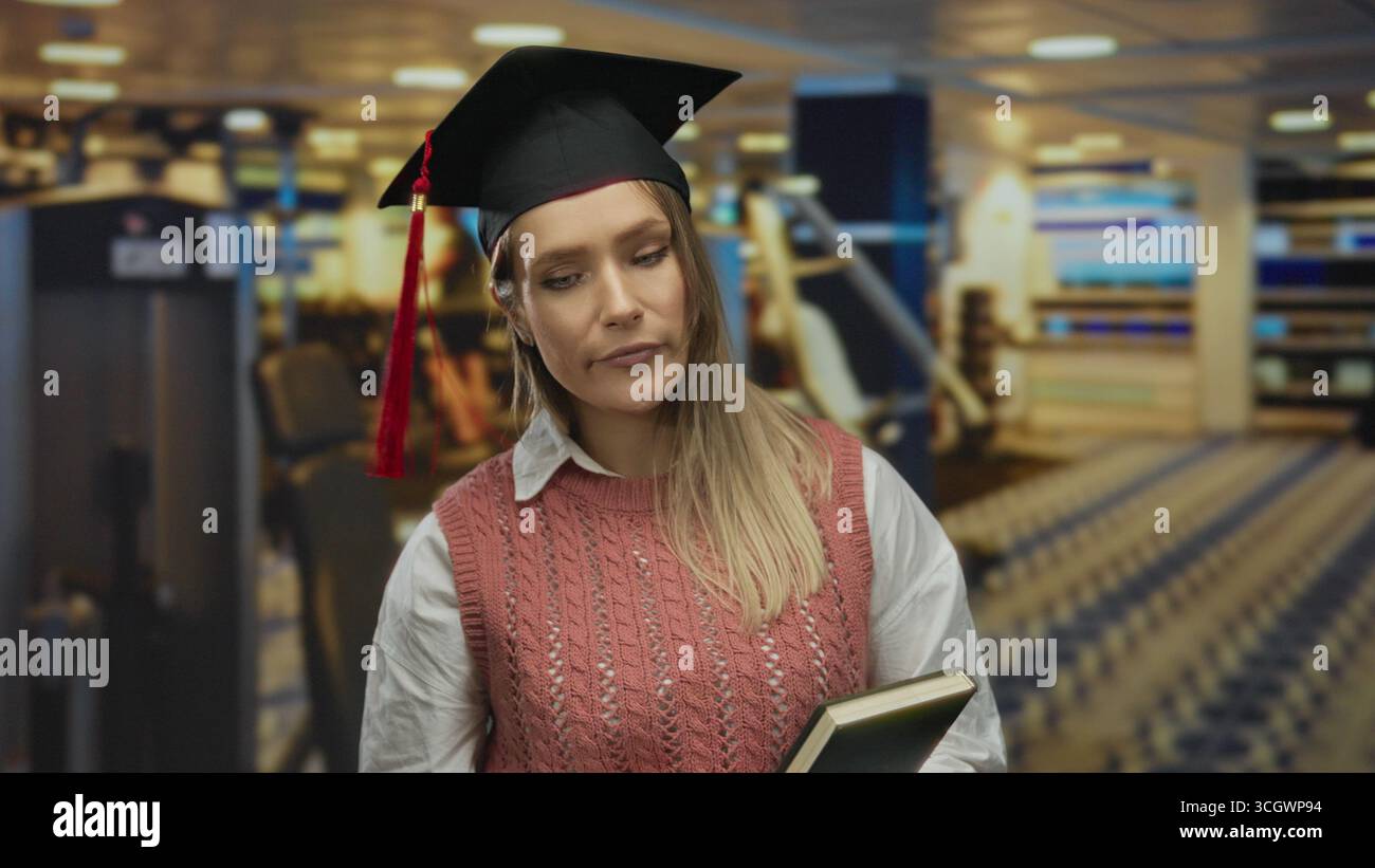 Woman in graduation cap holds book in gym, blending education with ...