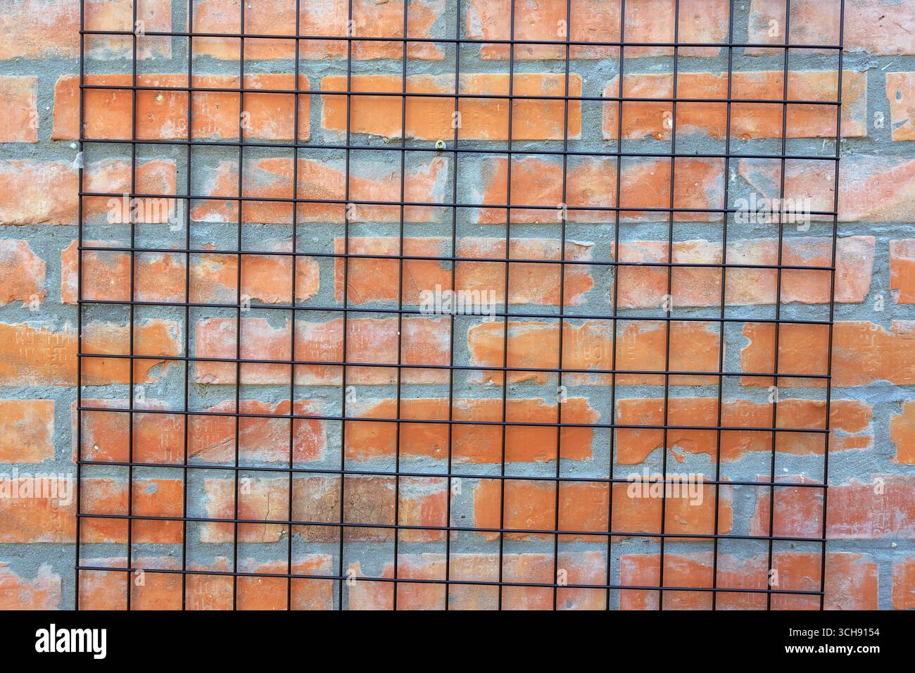 Red brick wall with visible mortar lines and a black steel wire mesh grid attached, texture ...