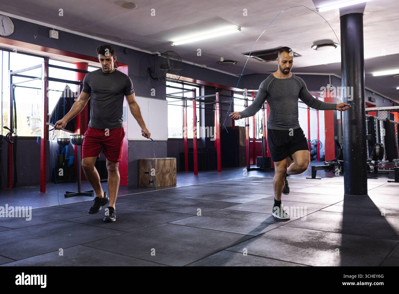 Diverse male workout partners jumping rope next to red squat racks on ...