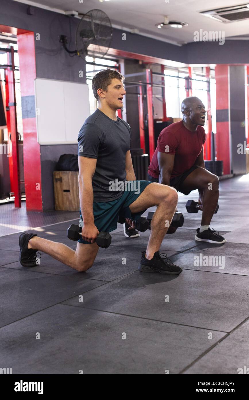 Male workout partners lunging forward with dumbbells in fitness center ...