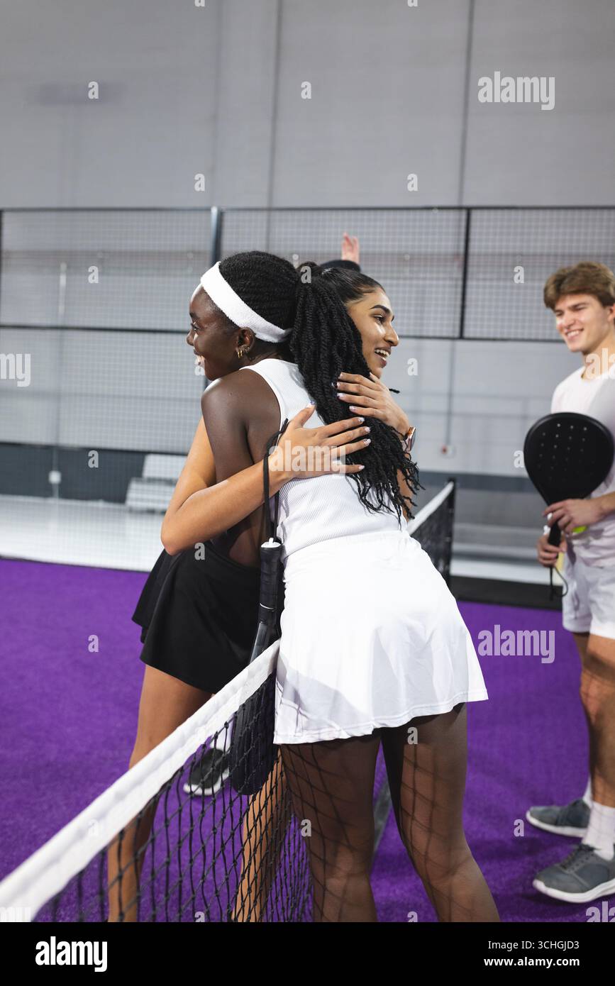 Diverse female tennis players hugging over net on court while male ...