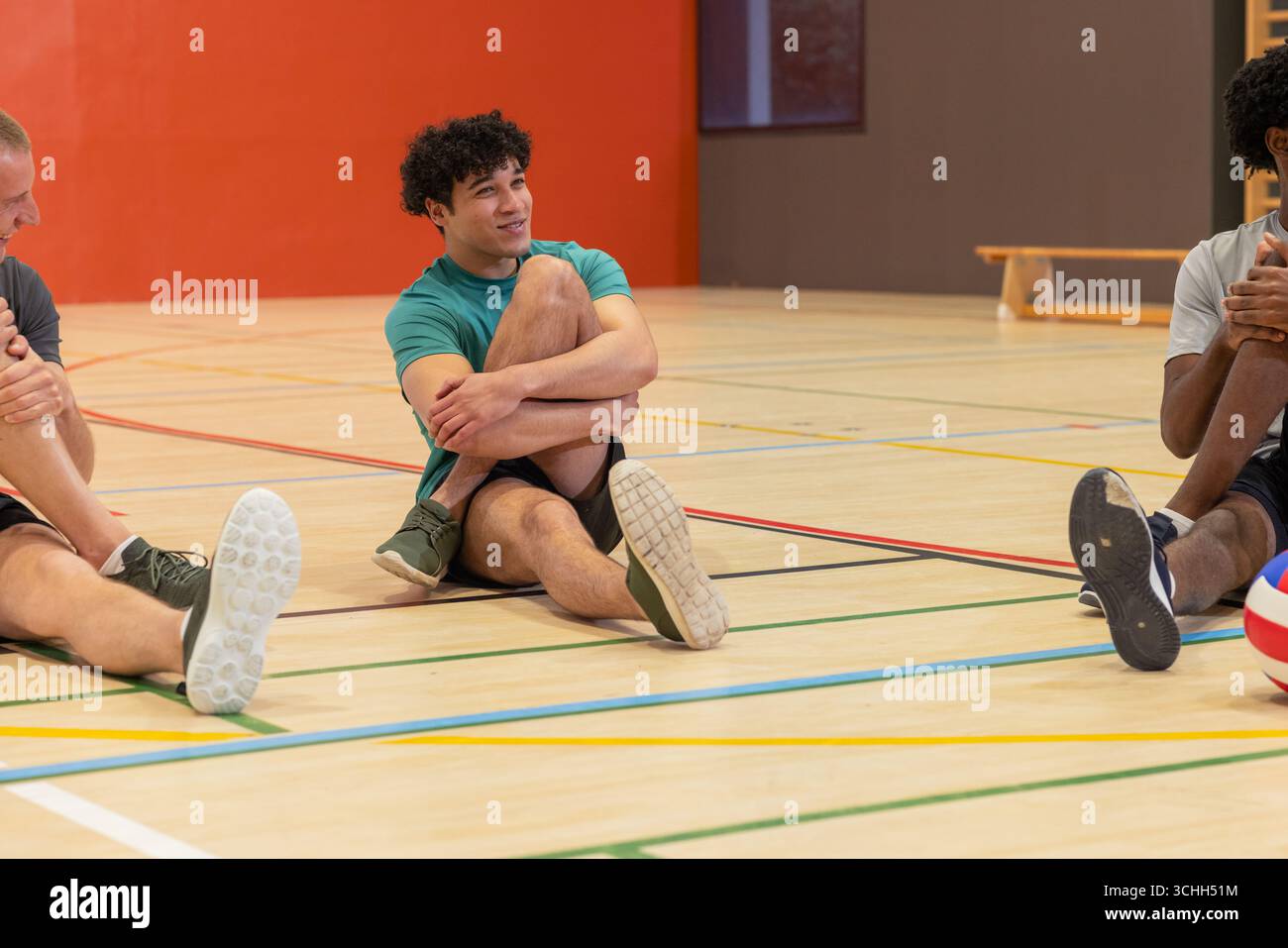 Diverse male teammates sitting on gym floor stretching legs by ...