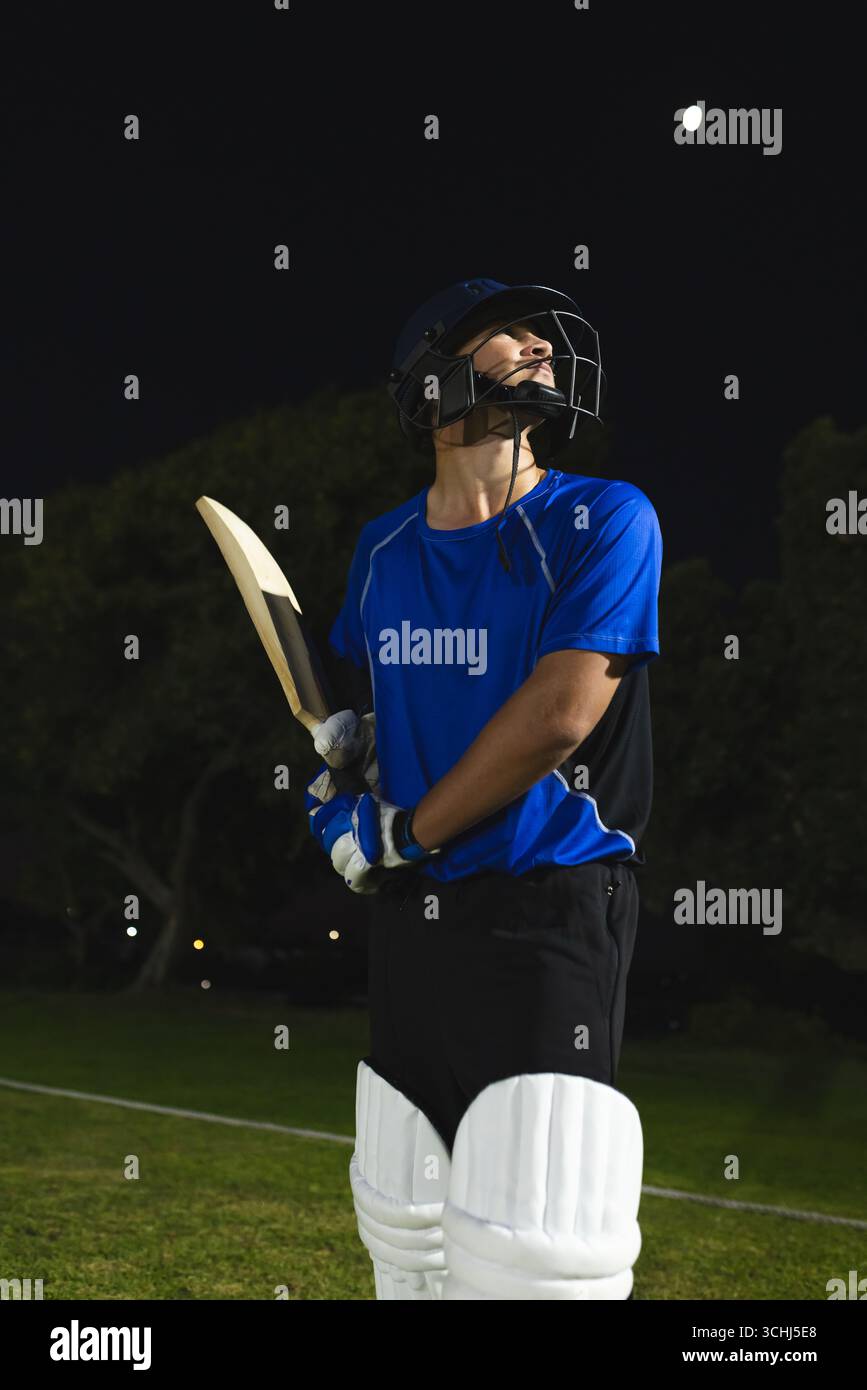 Male cricketer in helmet gripping cricket bat on grassy field at night ...