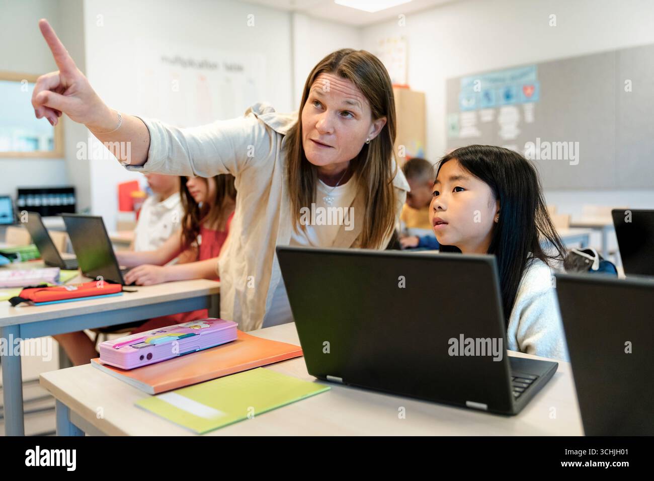 Female teacher pointing while standing next to girl sitting at desk in elementary classroom ...