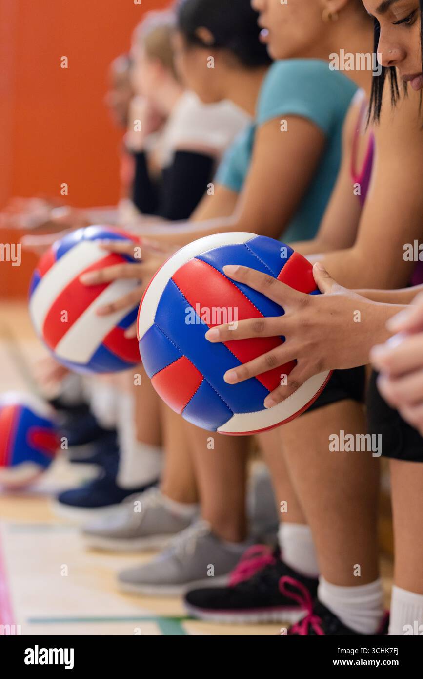 Diverse female volleyball teammates sitting on gym bench with red white ...