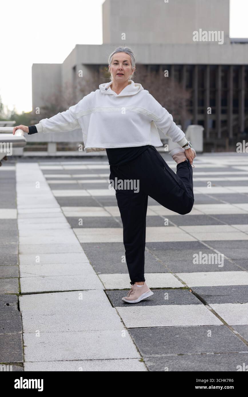 Senior woman stretching quadriceps with metal railing on tiled plaza ...
