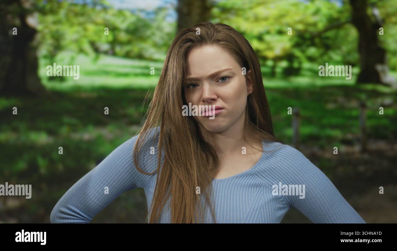 Woman frowns with a scrunched face and hands on hips in a sunlit park ...