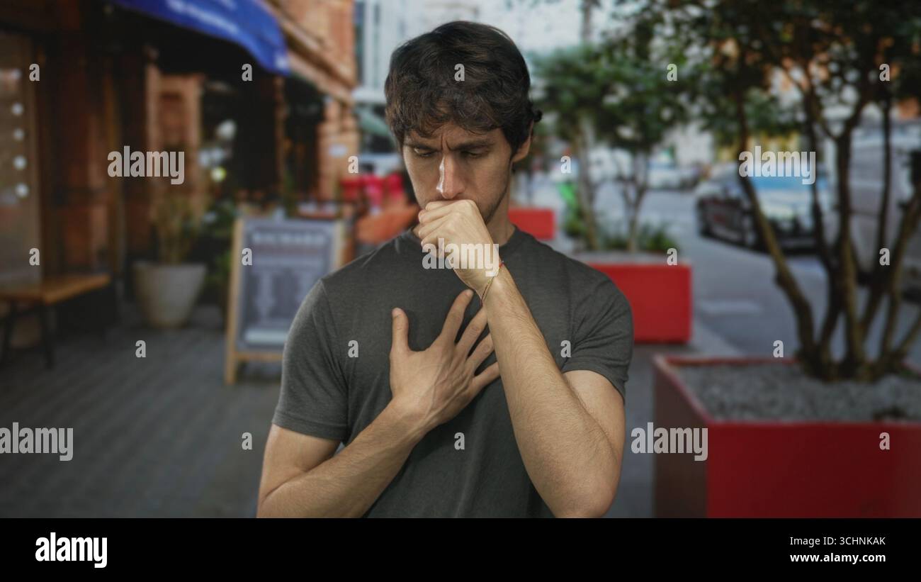 Man holding chest and coughing next to a red planter outside a sidewalk ...