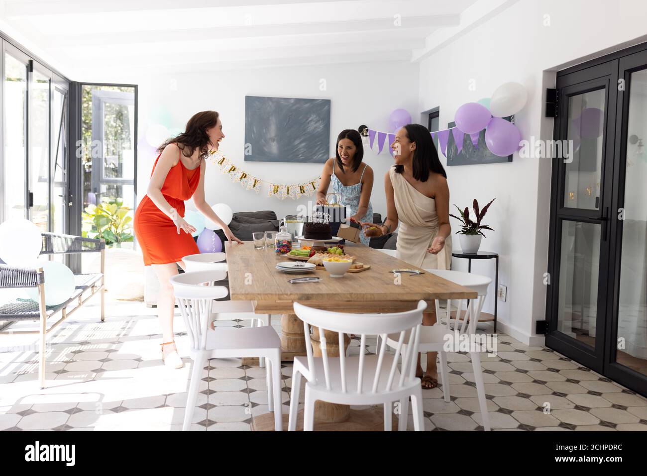 Setting up festive table, three women in their 30s laugh and chat ...