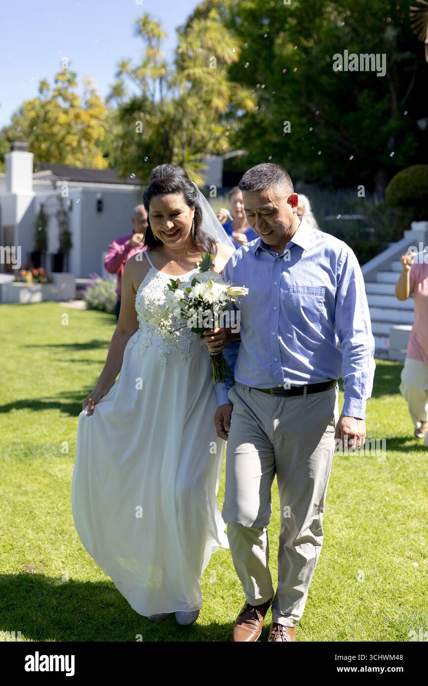 Walking with father, diverse bride smiling joyfully on sunny senior ...