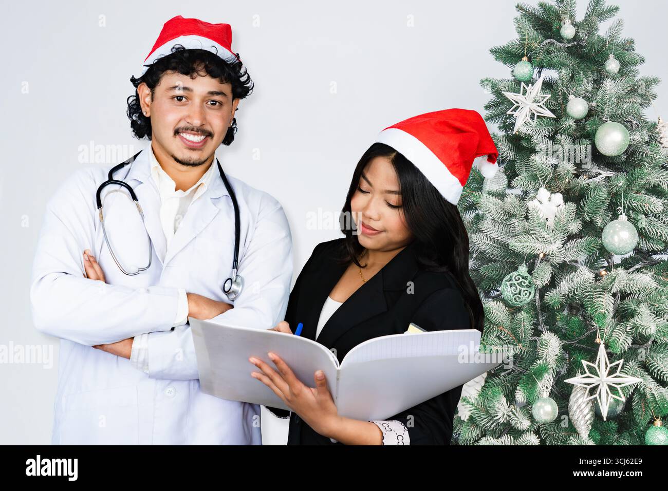 Doctor and nurse wearing Santa hats, standing next to a Christmas tree ...