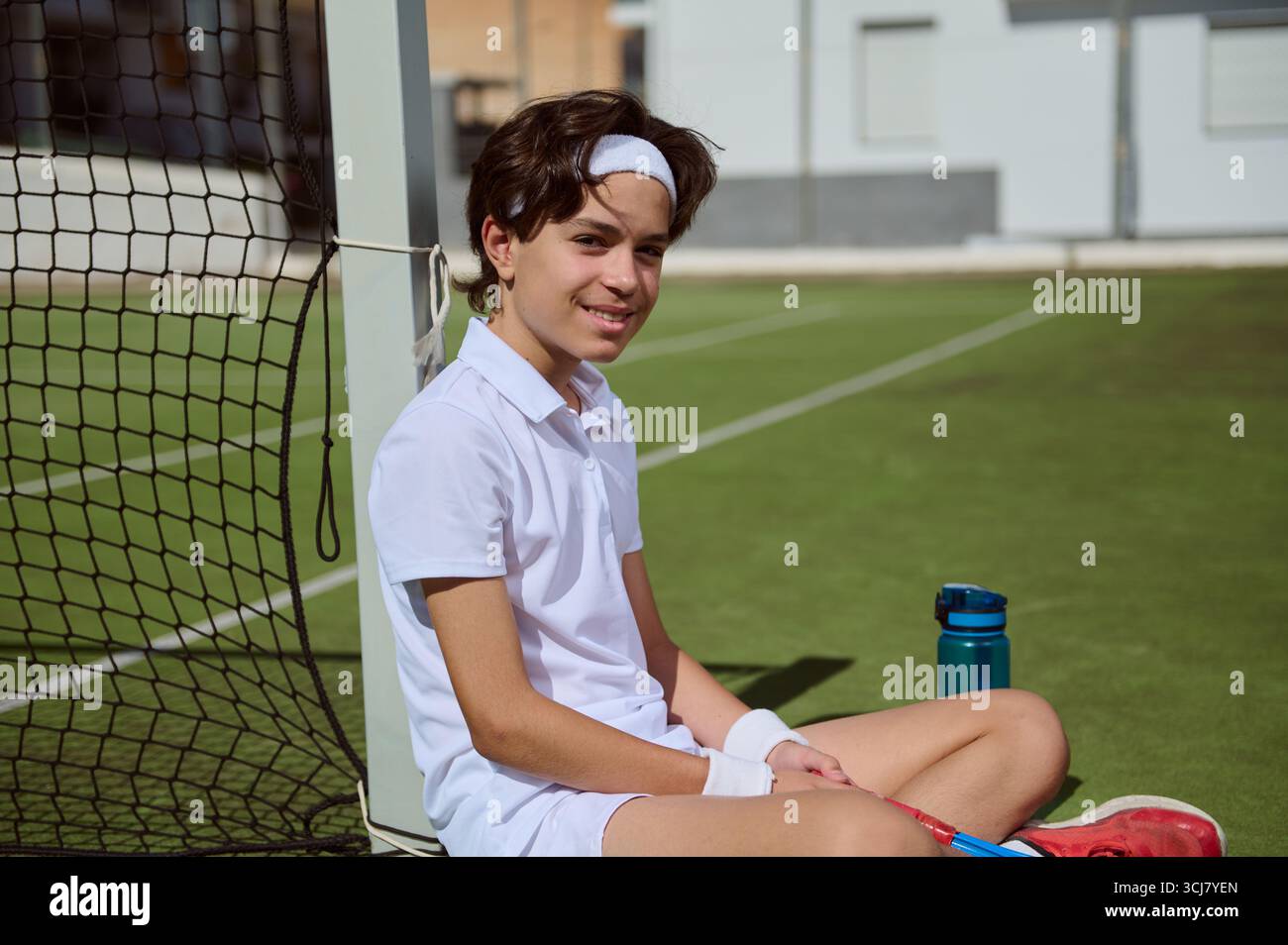 A young tennis athlete rests sitting on a tennis court after a session ...