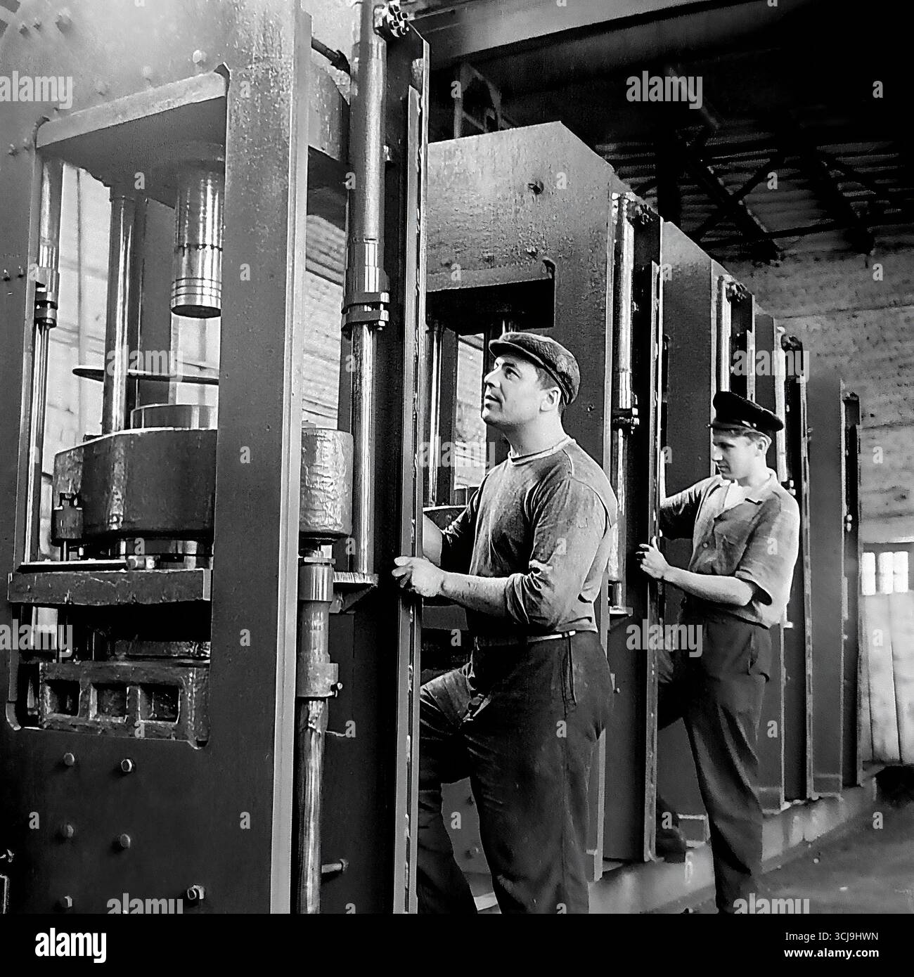 Two workers—a master and an apprentice—operate a giant hydraulic press ...
