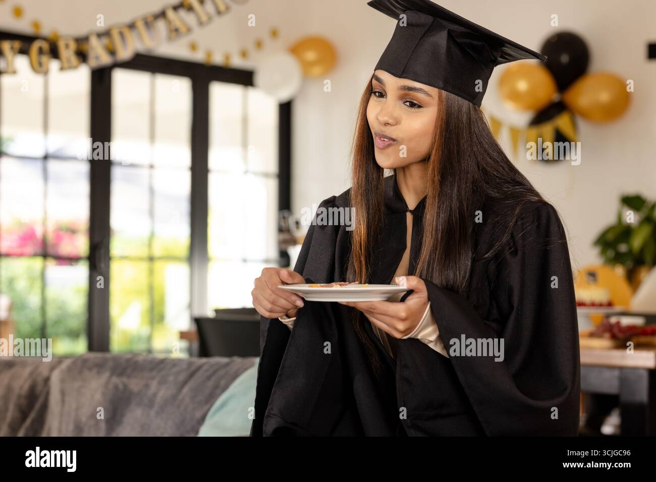 Graduate in cap and gown enjoying celebration at home with festive ...