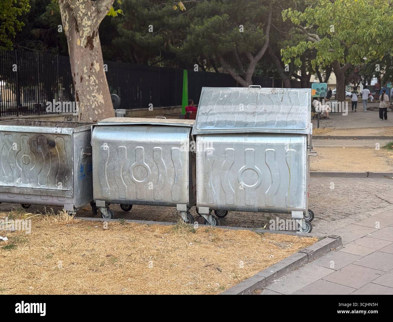 Metal garbage containers placed on pavement in a public area near trees ...