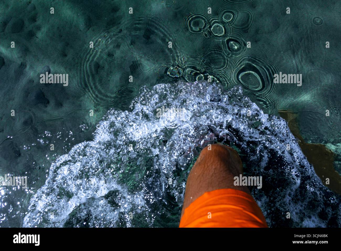 A man in orange swimming shorts walks in knee-deep water, blue water in ...
