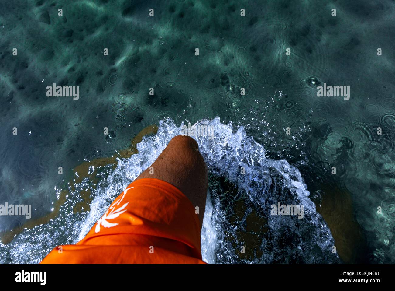 A man in orange swimming shorts walks in knee-deep water, blue water in ...