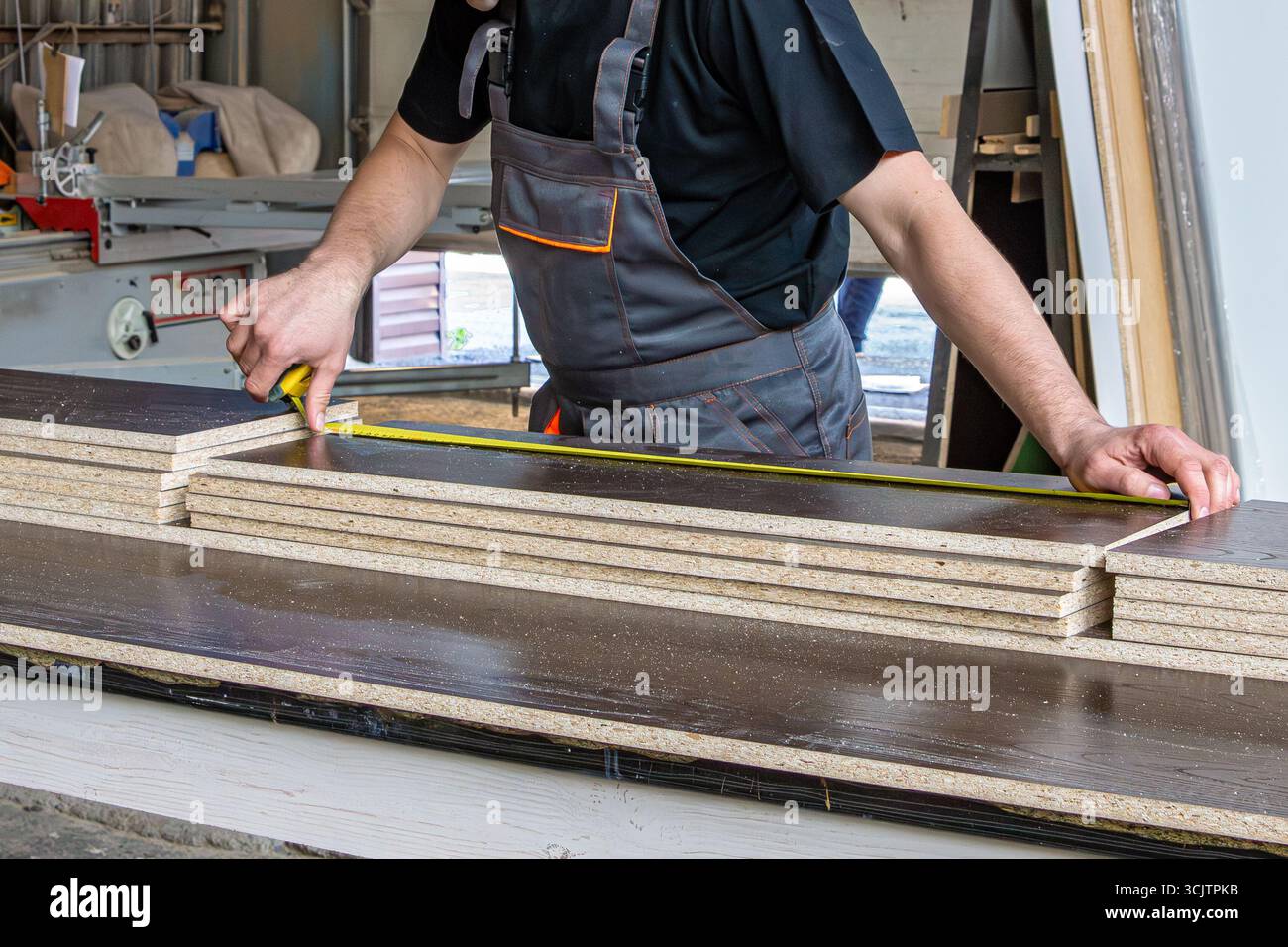 A skilled worker measures stacked laminated particleboard sheets in a workshop, with selective ...