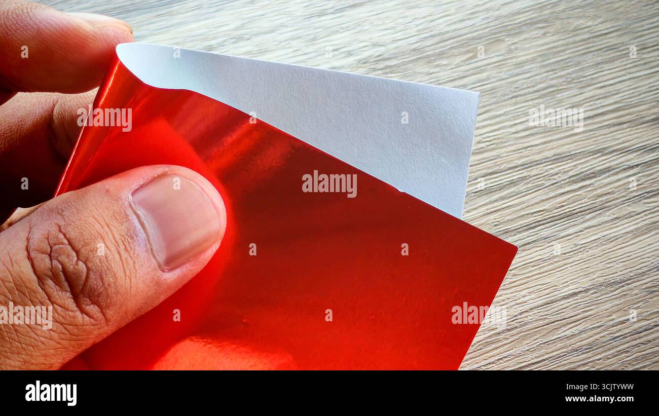 Hand peeling a metallic red origami paper sheet to reveal a white back ...