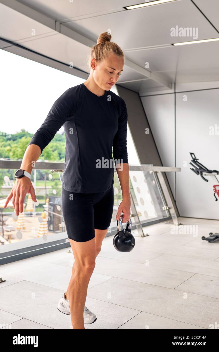 A focused woman dressed in black fitness attire performs kettlebell ...