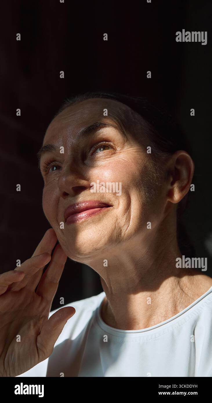 Elderly woman touching face with gentle smile under sunlight Stock ...