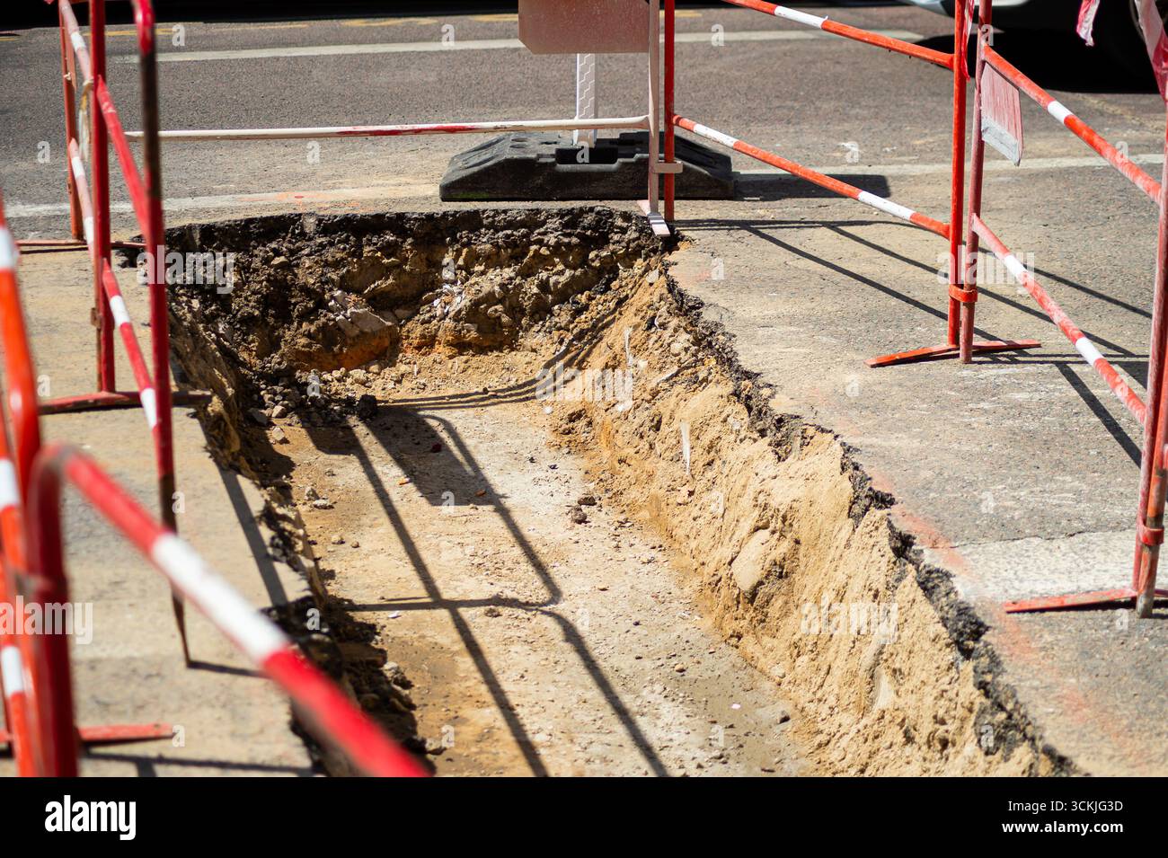 This photo shows an excavated trench on a road, surrounded by safety ...