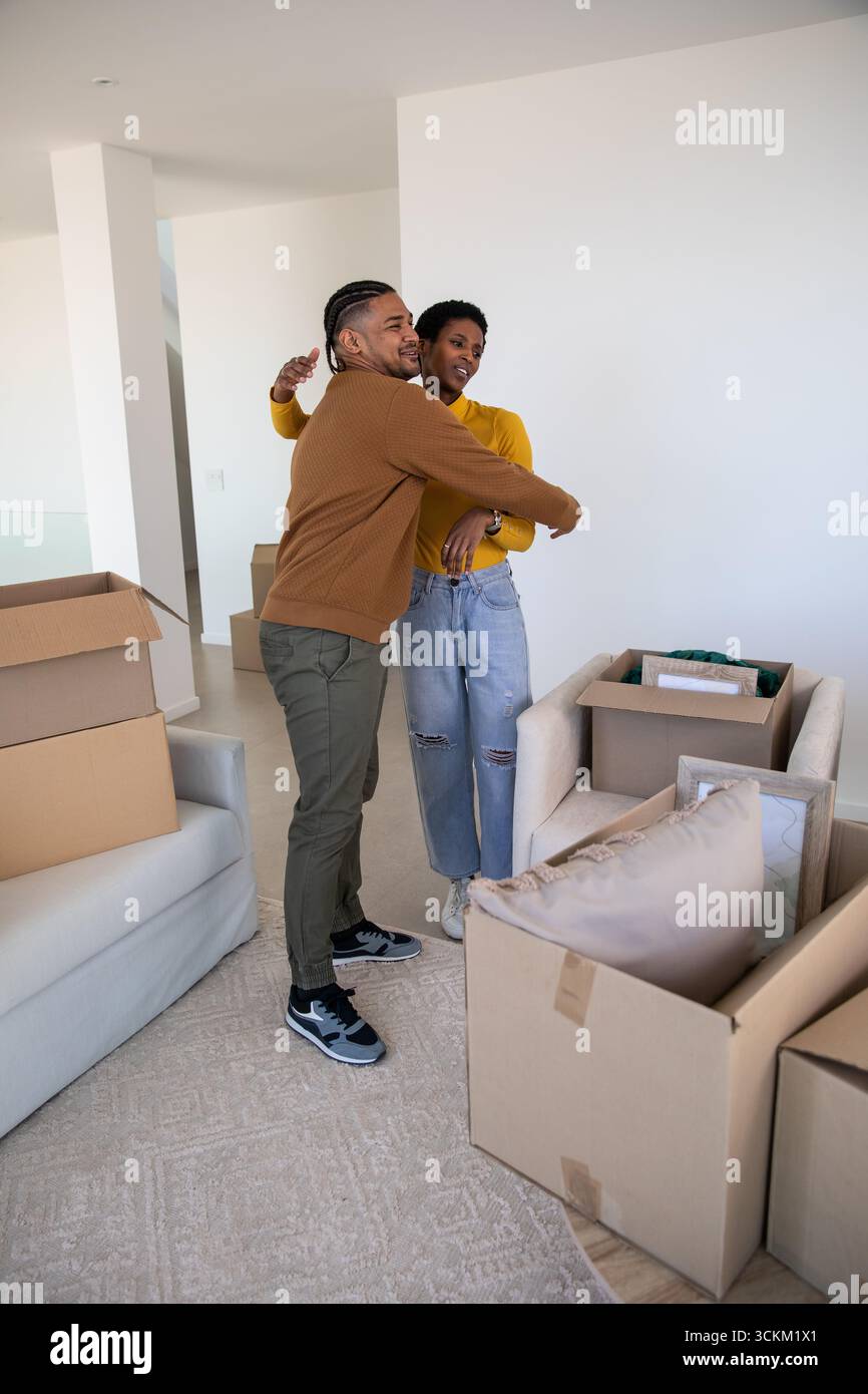 Diverse couple hugging in empty living room surrounded by moving boxes ...