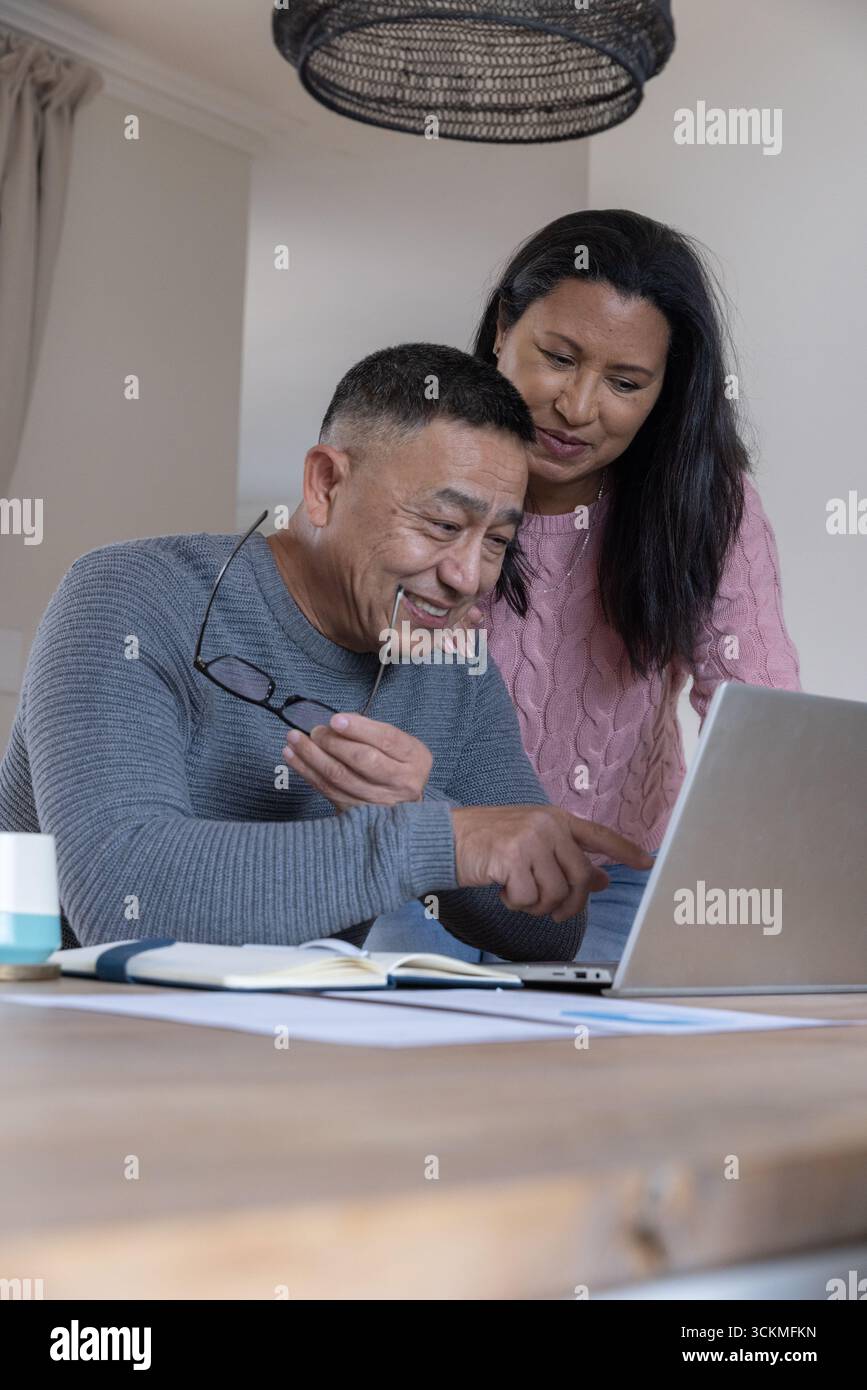 Diverse senior couple smiling, pointing at laptop at home on dining ...