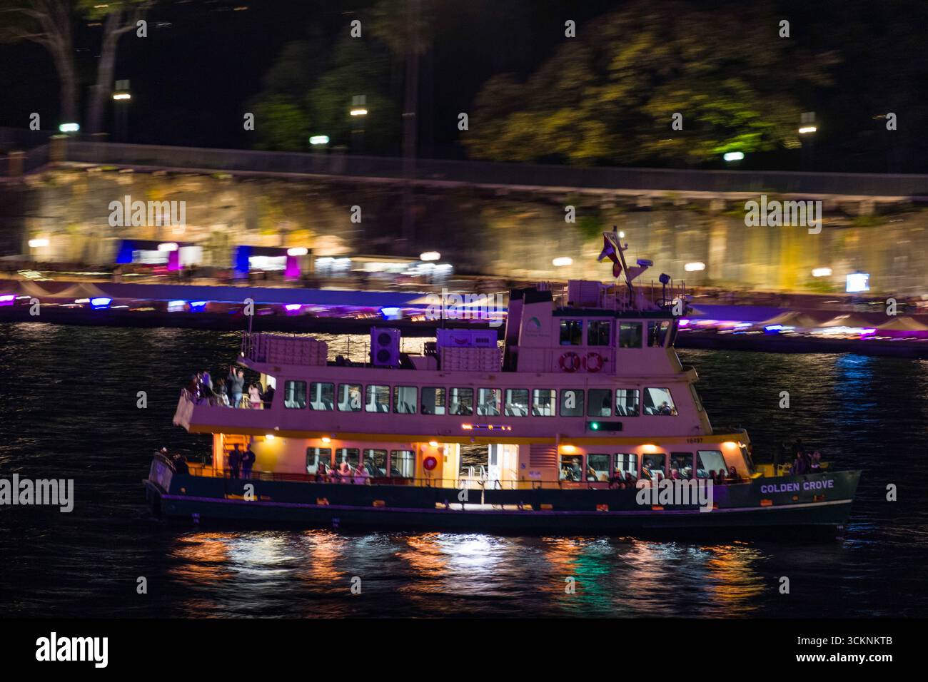 A ferry sails through a vibrant, illuminated city harbor at night, its ...